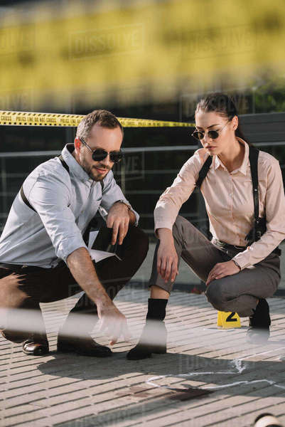 Male and female detectives sitting and looking at chalk line at crime ...