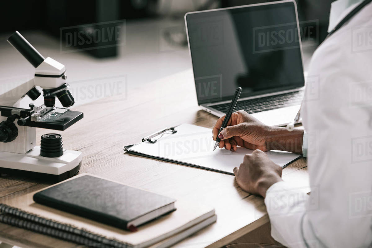 Cropped view of African American scientist writing on paper near laptop ...