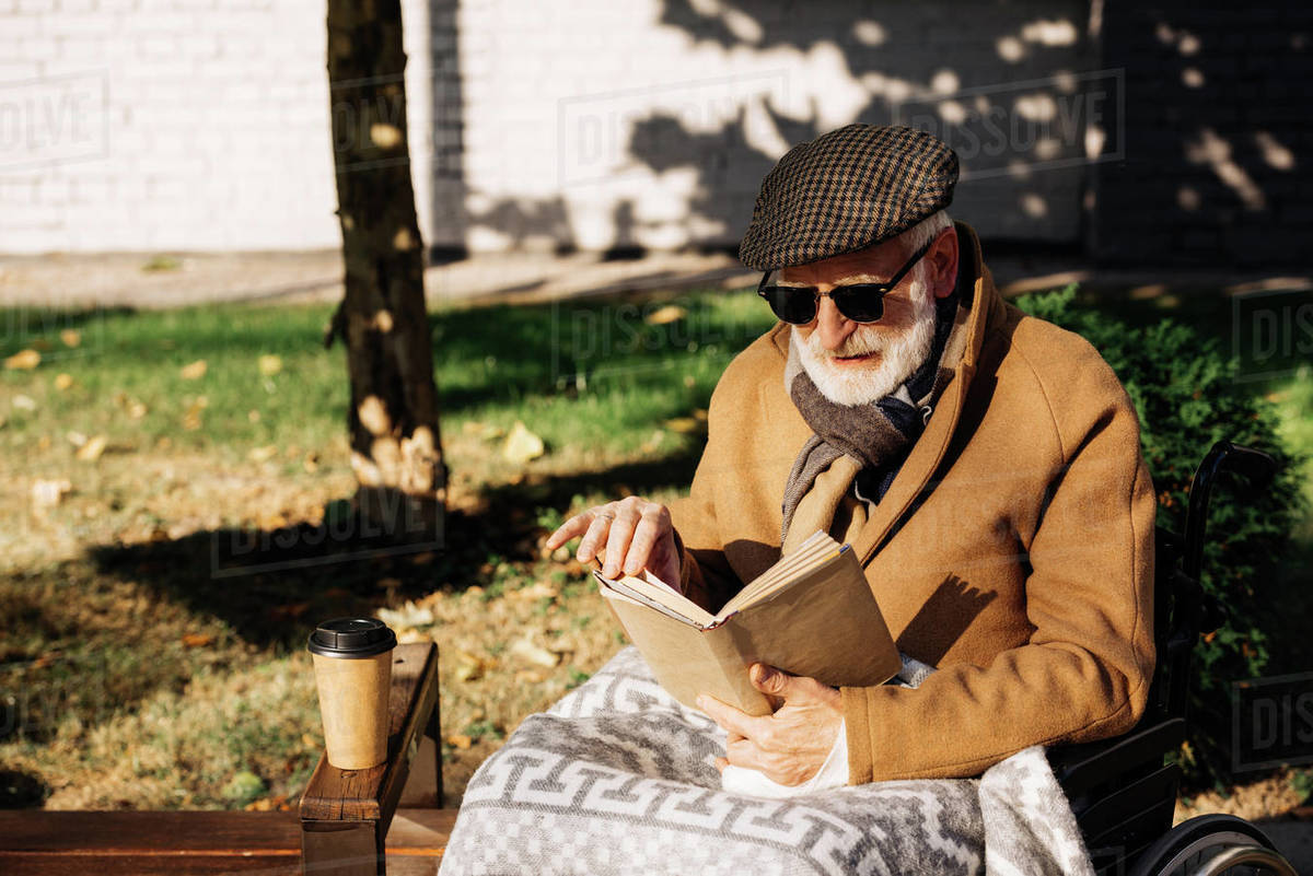 Handsome senior disabled man in wheelchair reading book on street ...