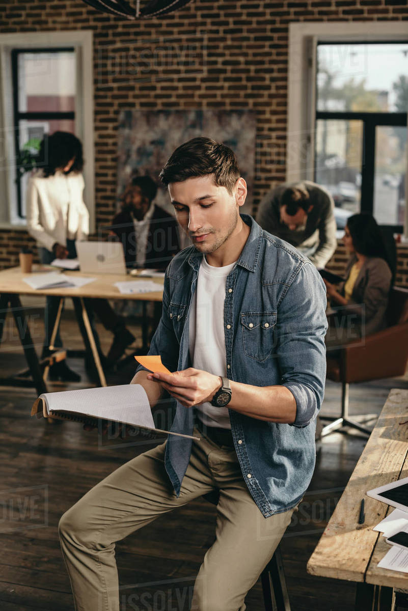 Young man holding journal and looking at note in modern loft office ...