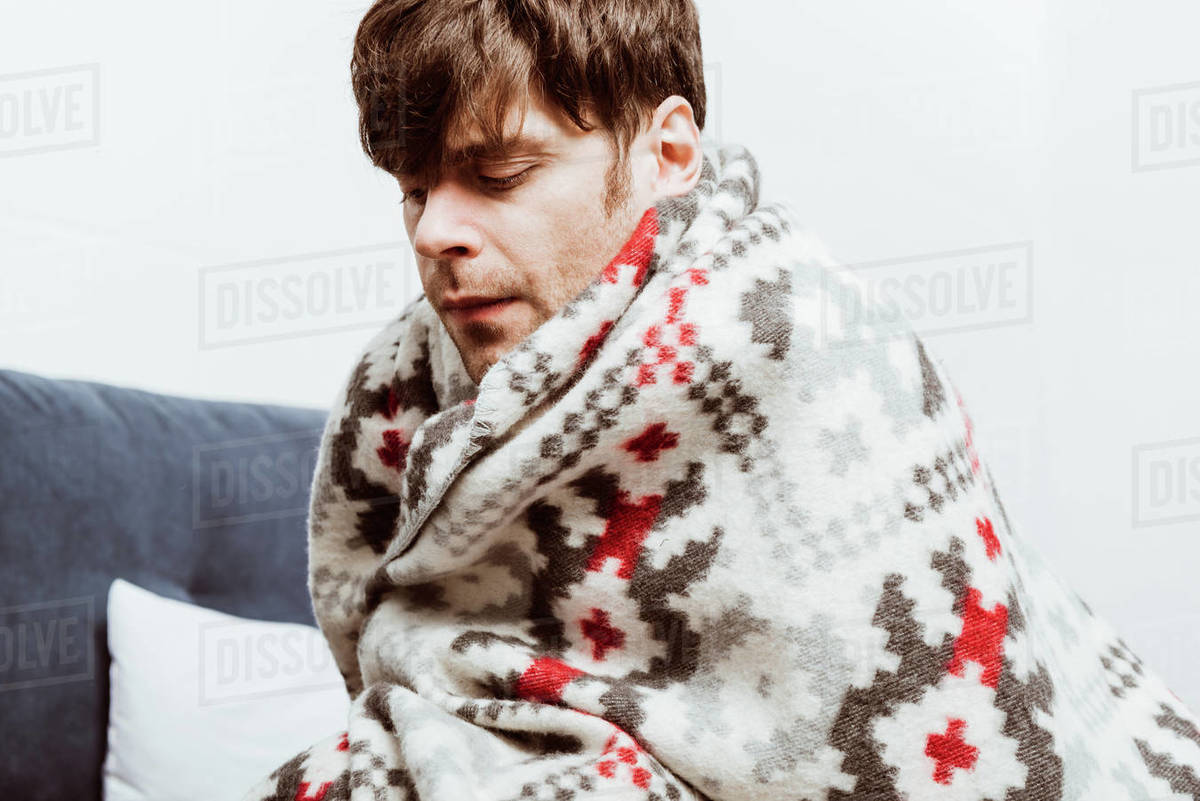 sick young man wrapped in blanket sitting on bed at home - Royalty-free ...