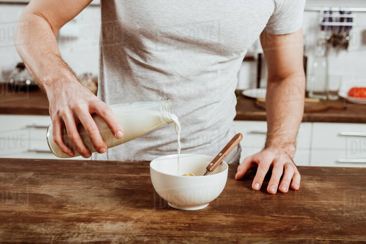 cropped image of man pouring milk into bowl with corn flakes in kitchen ...