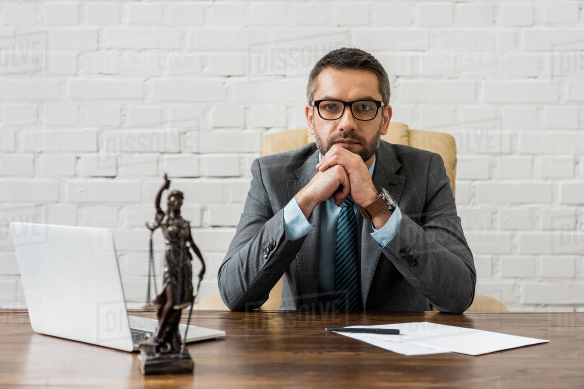 Serious bearded lawyer in suit sitting at workplace and looking at ...
