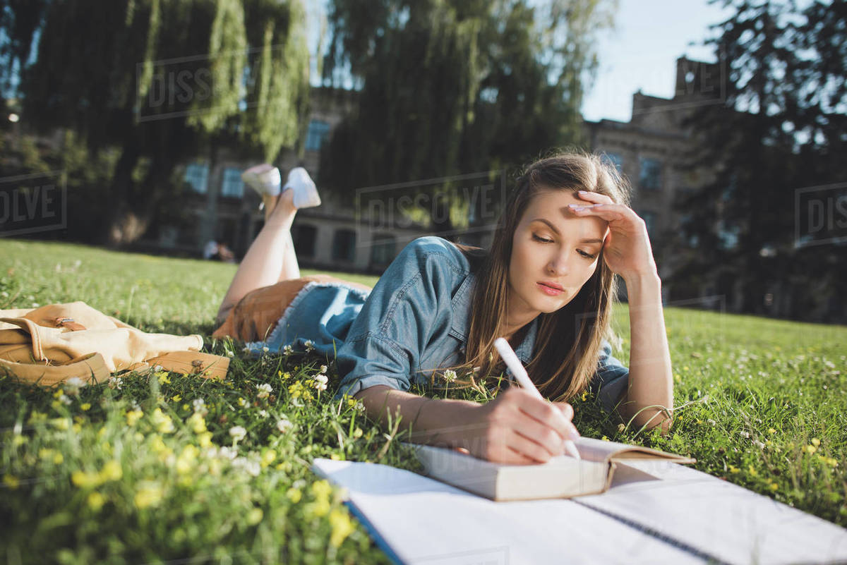Focused caucasian student doing homework while lying on grass in park ...