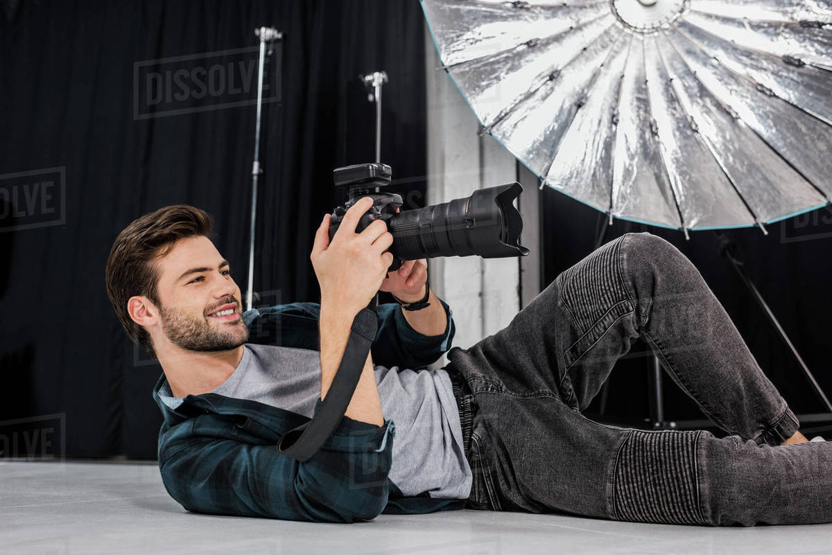 Smiling young photographer lying and using photo camera in studio ...