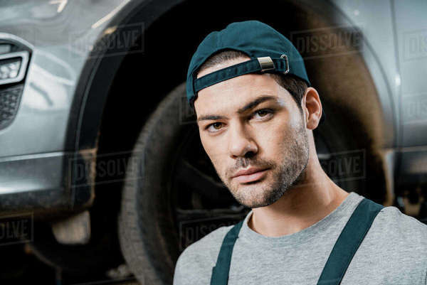 Portrait of young auto mechanic looking at camera with automobile ...