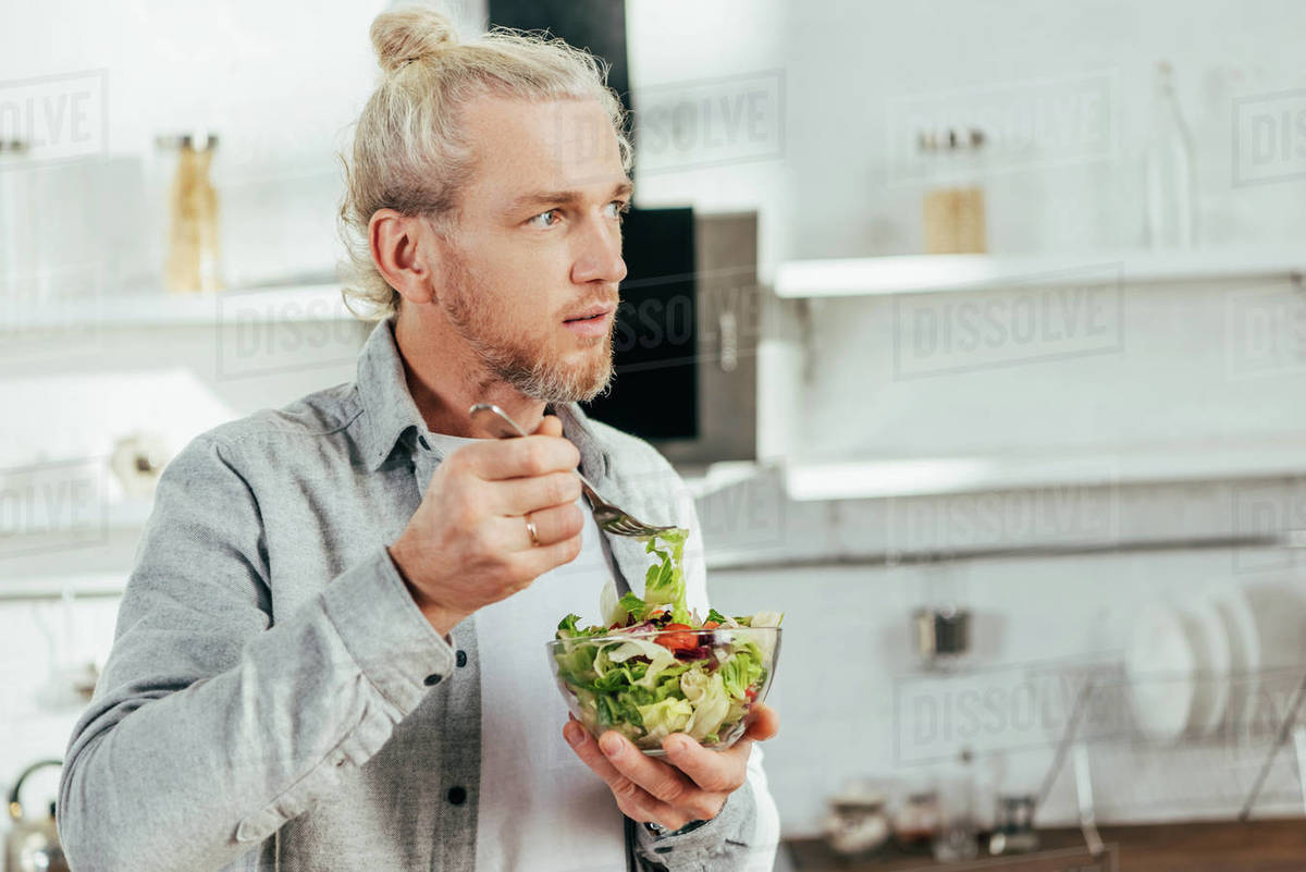 Handsome man eating vegetable salad and looking away in kitchen ...