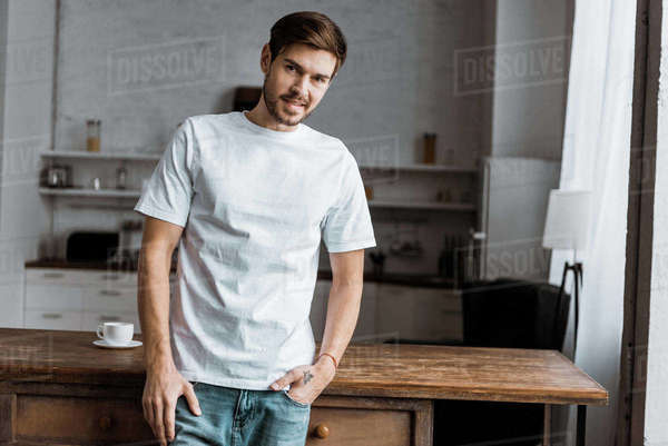 smiling young man leaning back on kitchen table and looking at camera ...