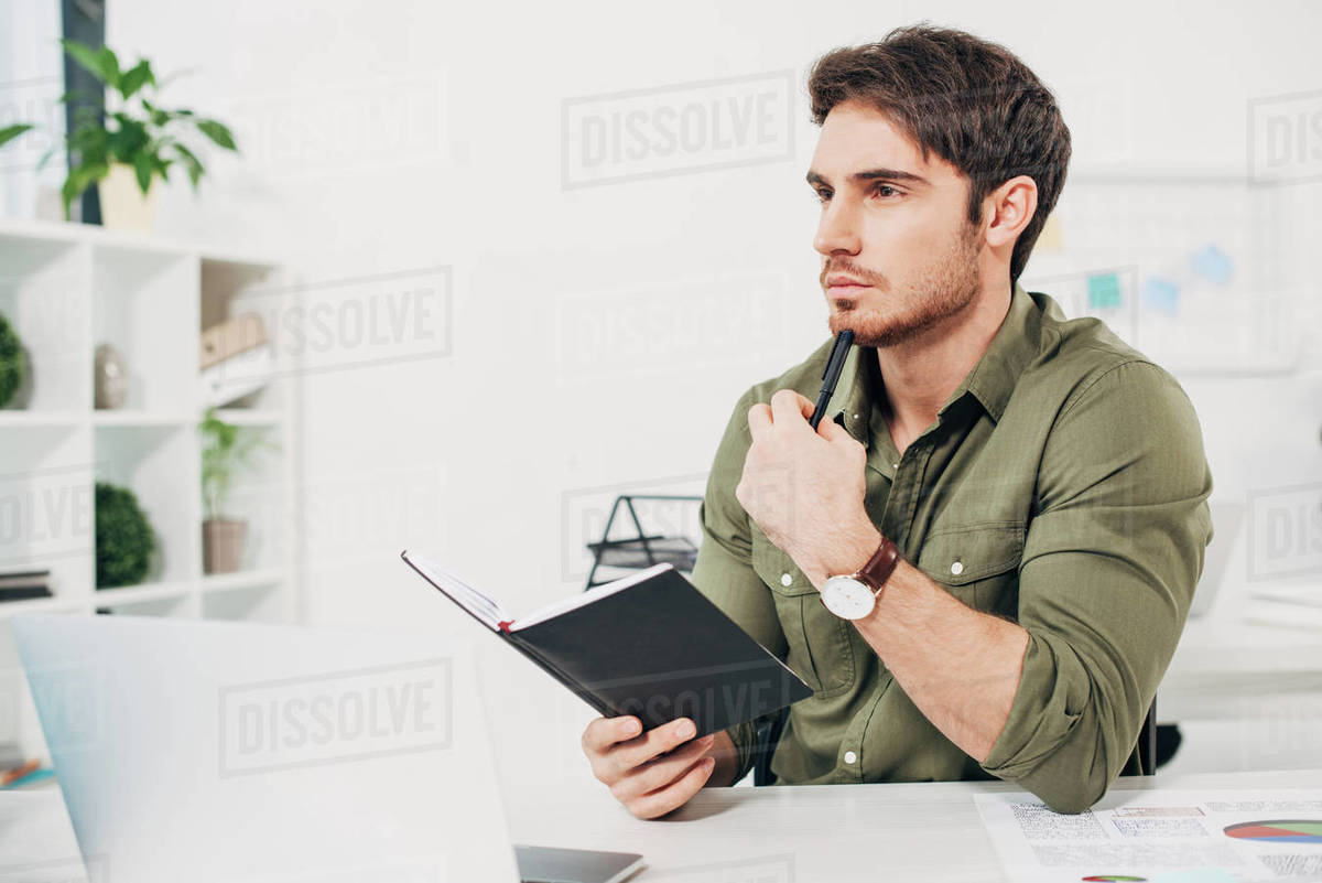 Pensive office manager sitting at desk and holding notebook and pen in ...
