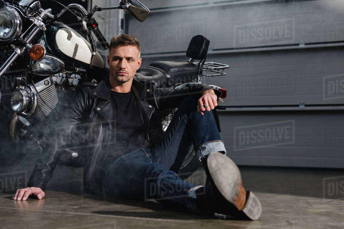 Handsome guy sitting on ground by motorcycle in garage - Royalty-free ...