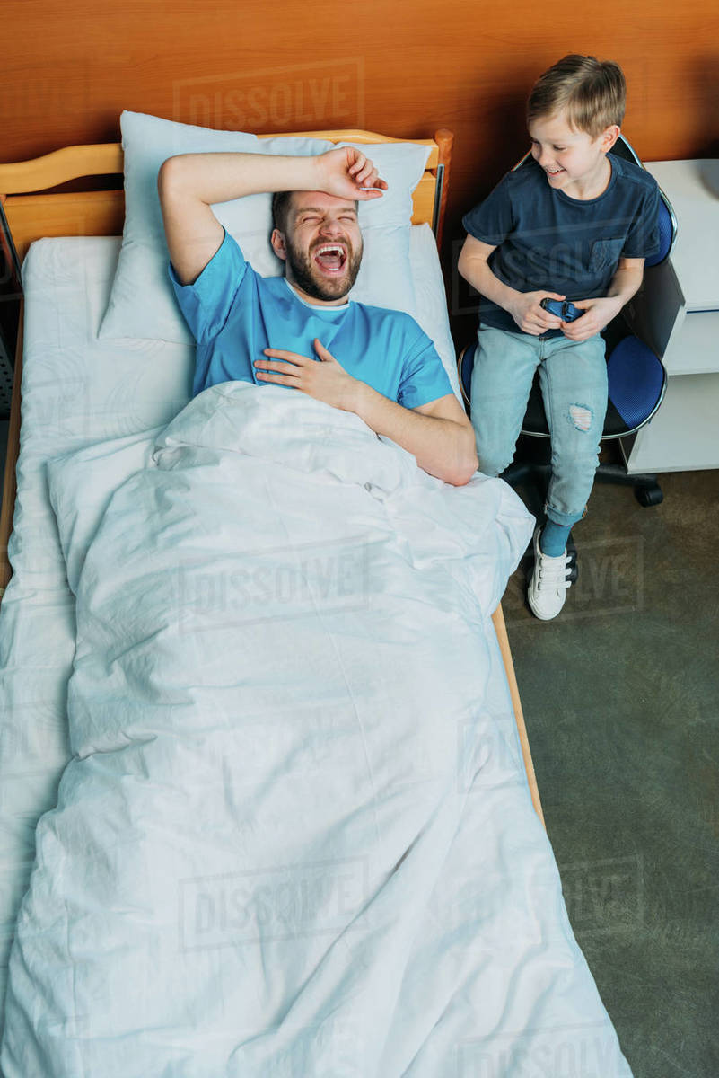 Son sitting near his sick father laying on hospital bed at ward ...