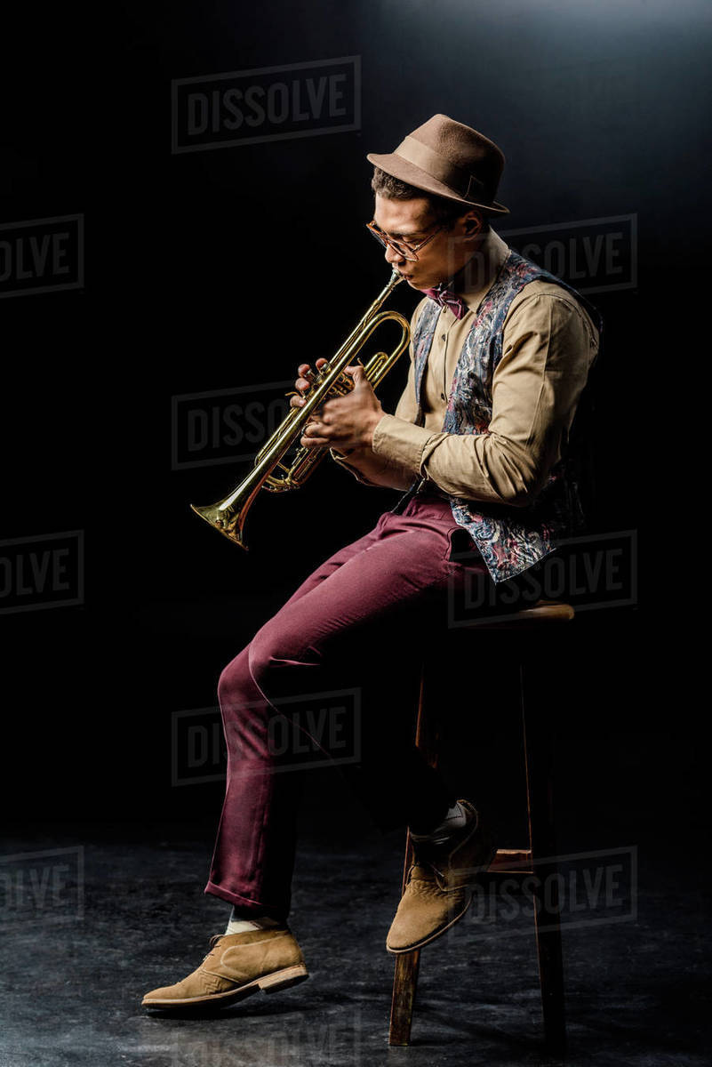 Stylish mixed race male musician playing on trumpet while sitting on ...