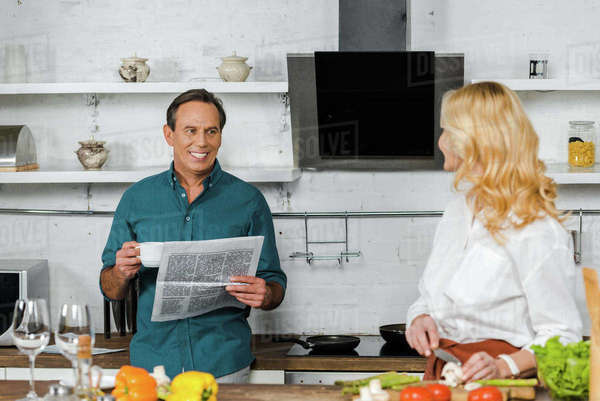 Mature wife cooking and smiling husband reading newspaper in kitchen ...