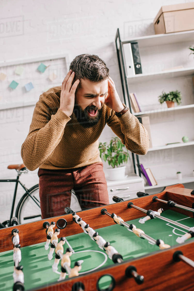 Angry adult man with hands on head yelling and playing table football ...