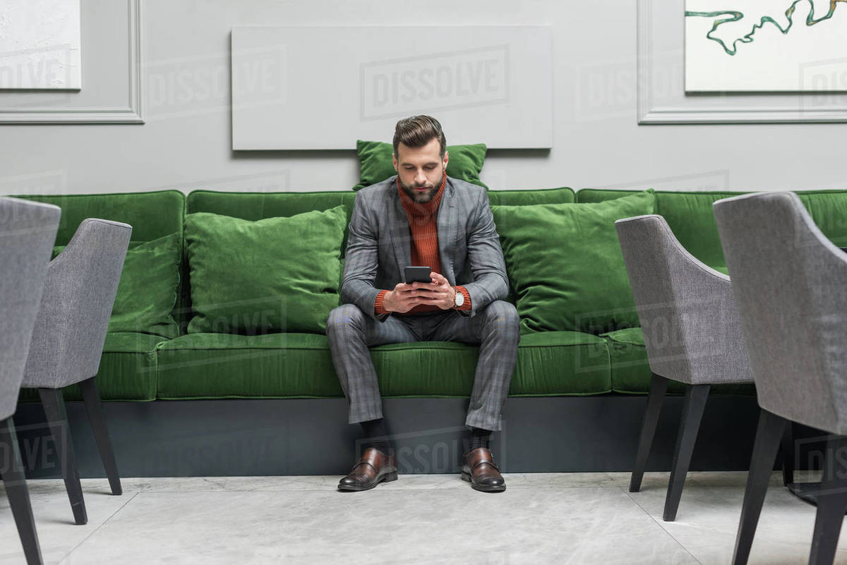 Serious handsome man in formal wear sitting on green sofa and using ...