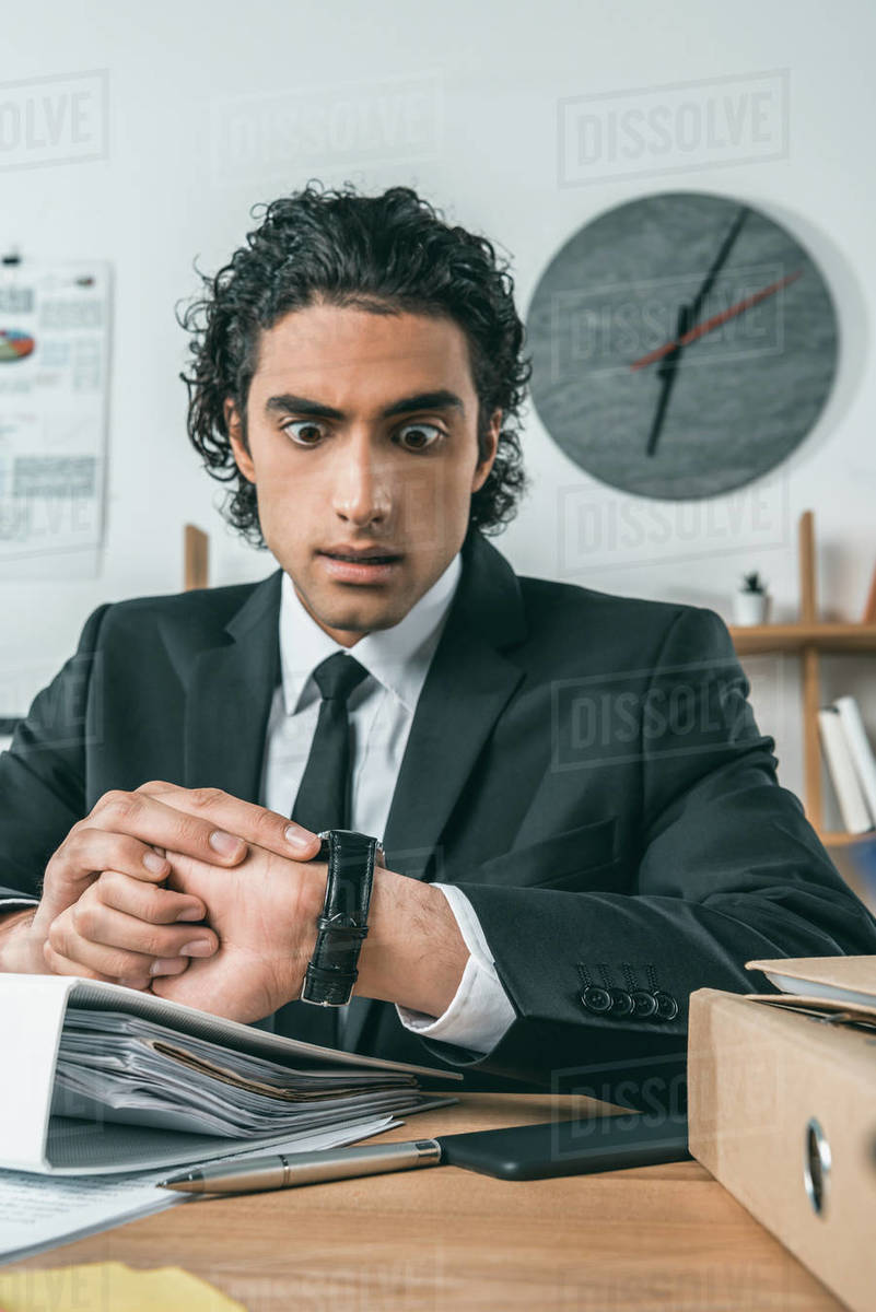 Portrait of surprised businessman checking time on watch while sitting ...