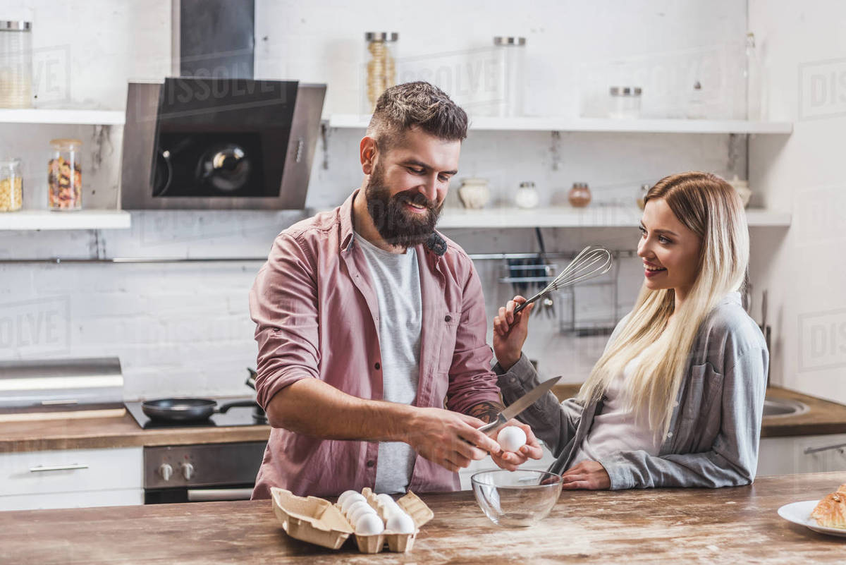 Handsome bearded man smashing eggs at wooden table while woman holding ...
