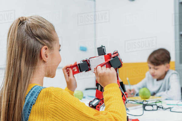 Schoolgirl sitting at table and holding robot model during stem lesson ...