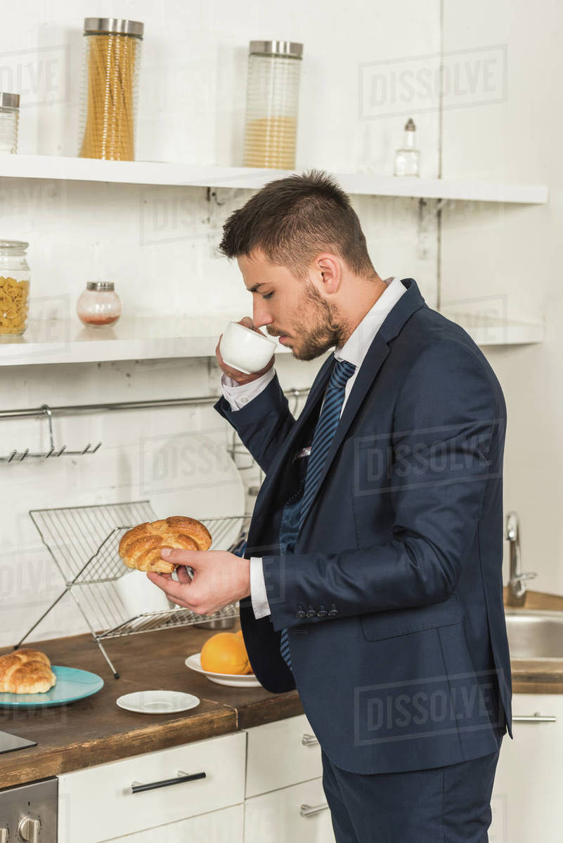 Side view of handsome man in suit drinking coffee and holding croissant ...