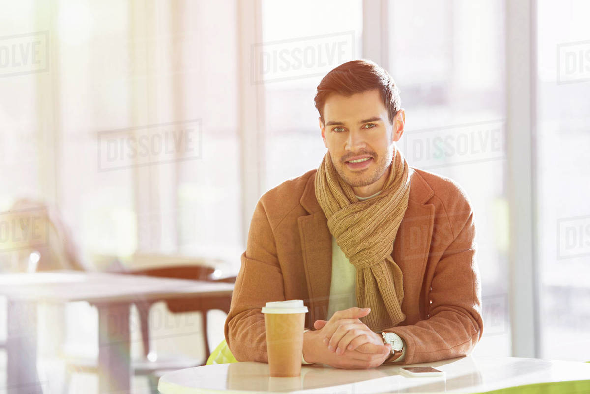 Handsome man smiling and sitting at table with disposable cup in cafe ...