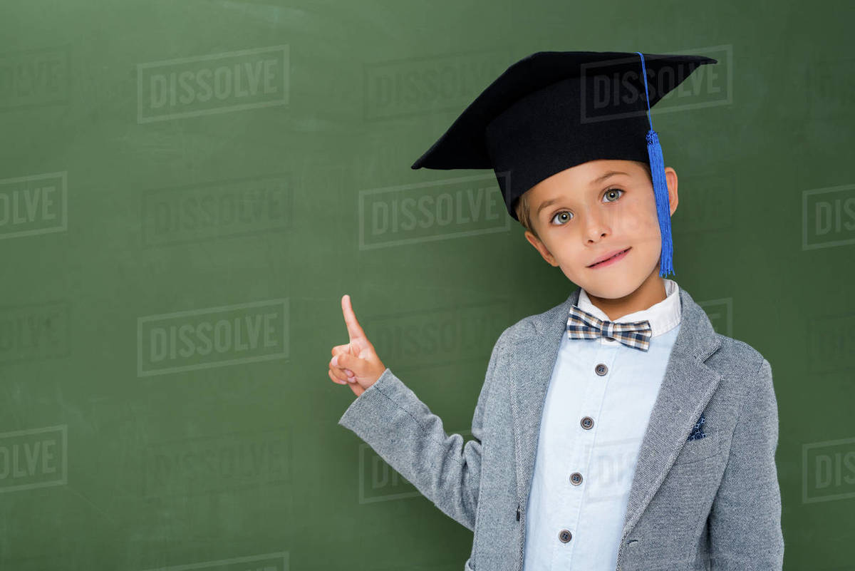 Schoolboy in graduation hat pointing at copy space next to chalkboard ...