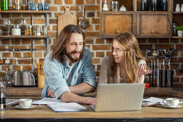 Attractive smiling couple talking and gesturing while using laptop at ...