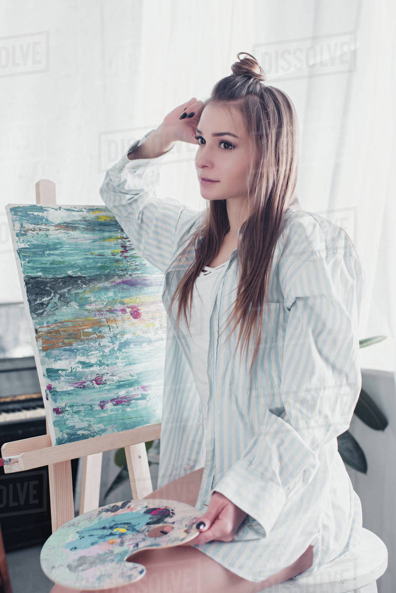 Thoughtful artist sitting on chair in front of easel and holding ...
