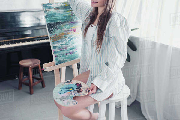 Cropped view of artist sitting on chair in front of easel and holding ...