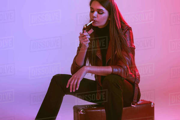 stylish smoking model in leather jacket sitting on suitcase, on pink ...