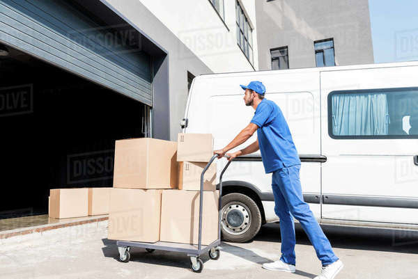 Young delivery man with cardboard boxes on cart driving into warehouse ...
