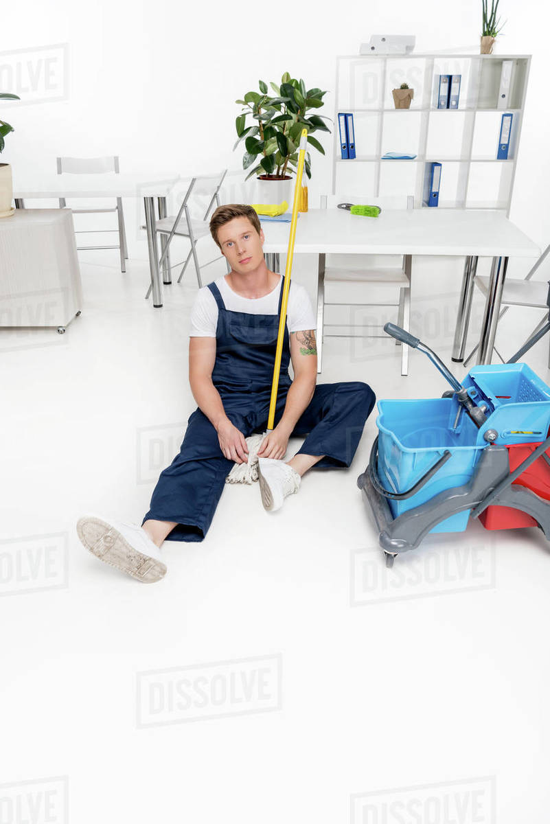 handsome young cleaner with mop sitting on floor and looking at camera ...