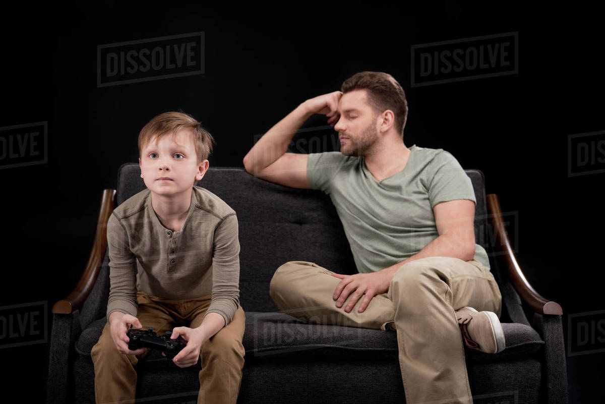 Upset father sitting on sofa near little son playing with joystick ...