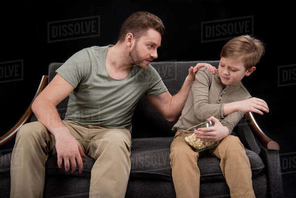 Angry father looking at scared little son sitting on sofa with popcorn ...