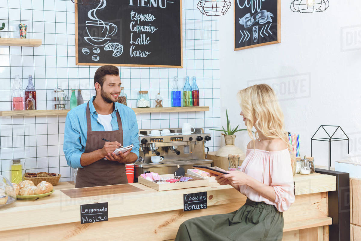 Handsome smiling barista taking notes and looking at young woman with ...