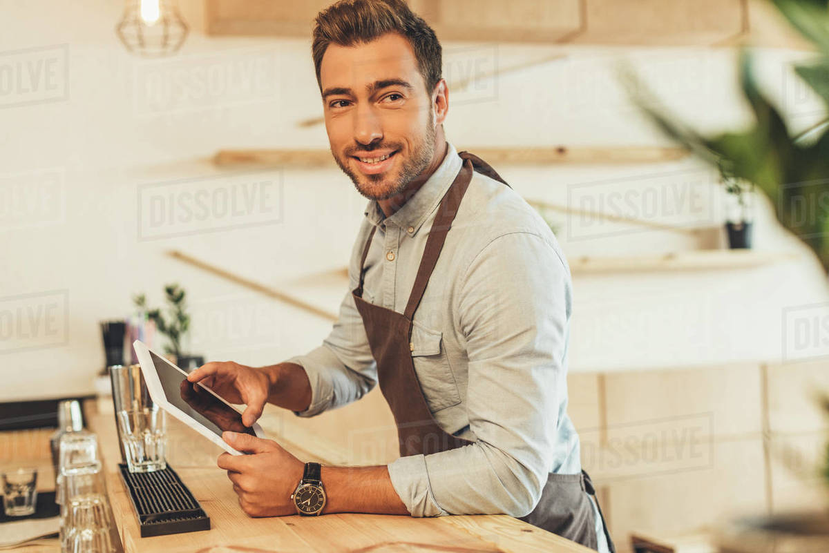 Side view of smiling barista with tablet looking away in coffee shop ...