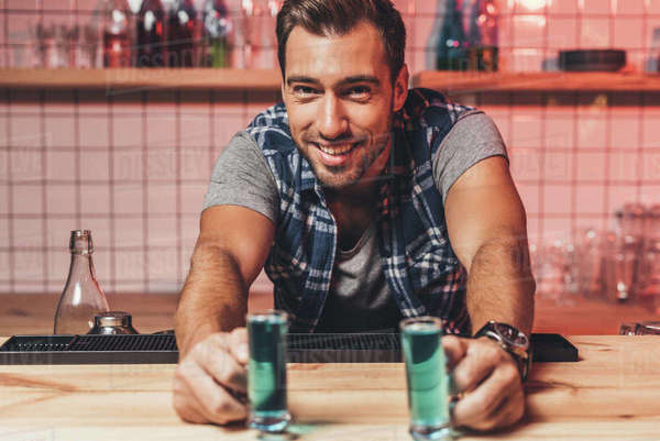 Portrait of cheerful barman with alcohol shots looking at camera in bar ...