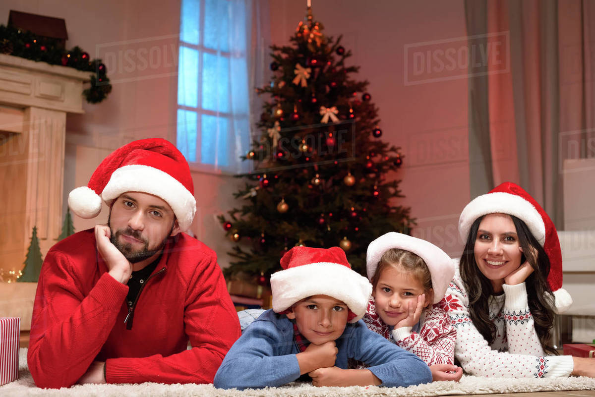 Happy family in santa hats lying together on carpet and looking at ...