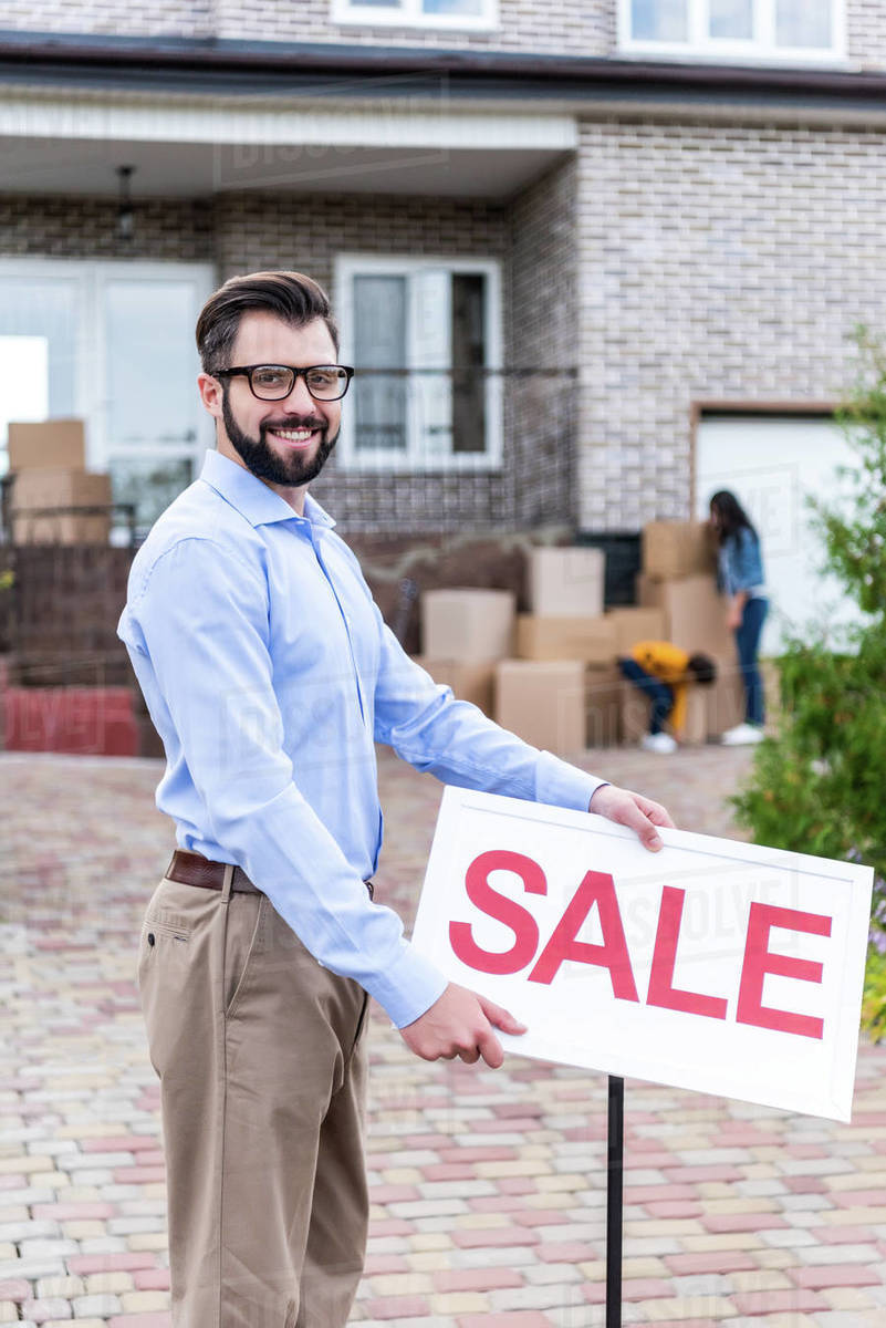 Young realtor with sale signboard in front of house with people moving ...