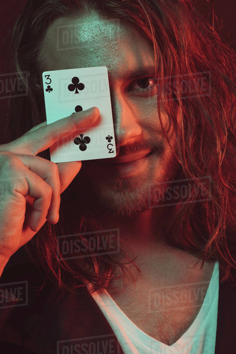 Portrait of young smiling man holding playing card in front of face ...