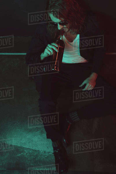 Young boozer in sunglasses drinking alcohol from bottle while sitting ...
