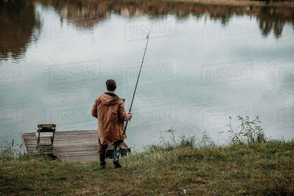 Rear view of fisherman fishing with rod on wooden pier at lake ...