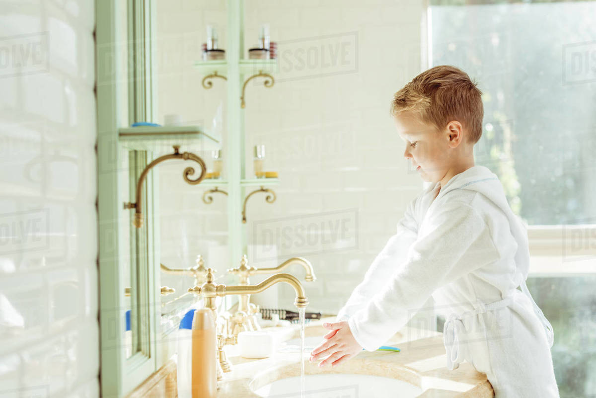 Side view of cute little boy in bathrobe washing hands in bathroom ...