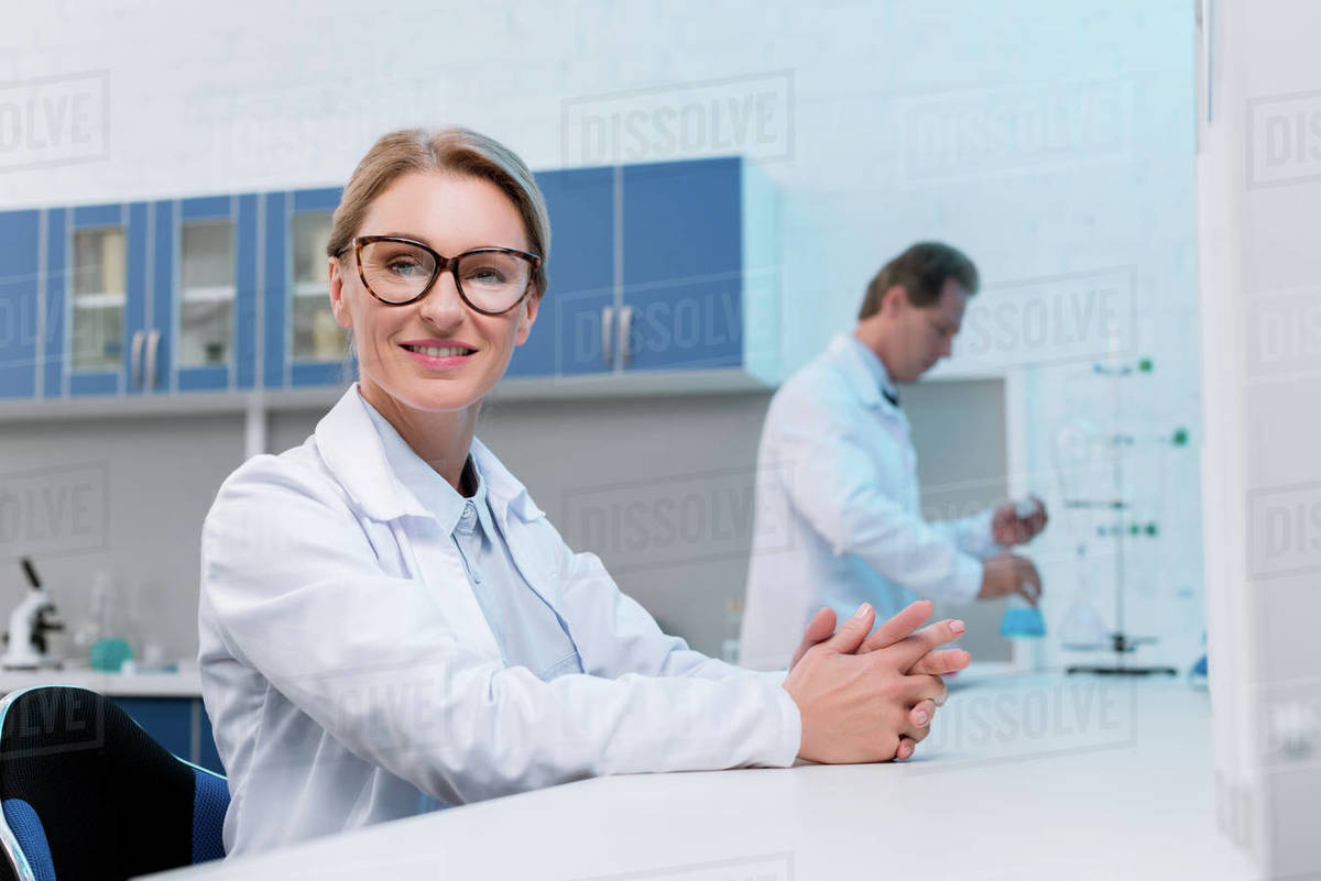 Beautiful chemist in eyeglasses sitting at table and smiling at camera ...