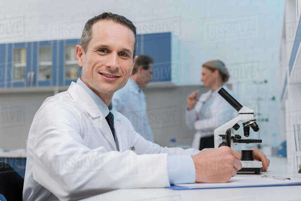 Smiling lab technician taking notes while doing microscope sample ...