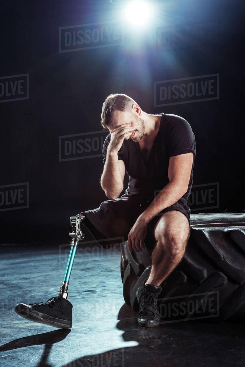 Sad sportsman with leg prosthesis sitting on tire after training ...