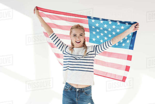 Cheerful blonde girl holding us flag and smiling at camera on grey ...
