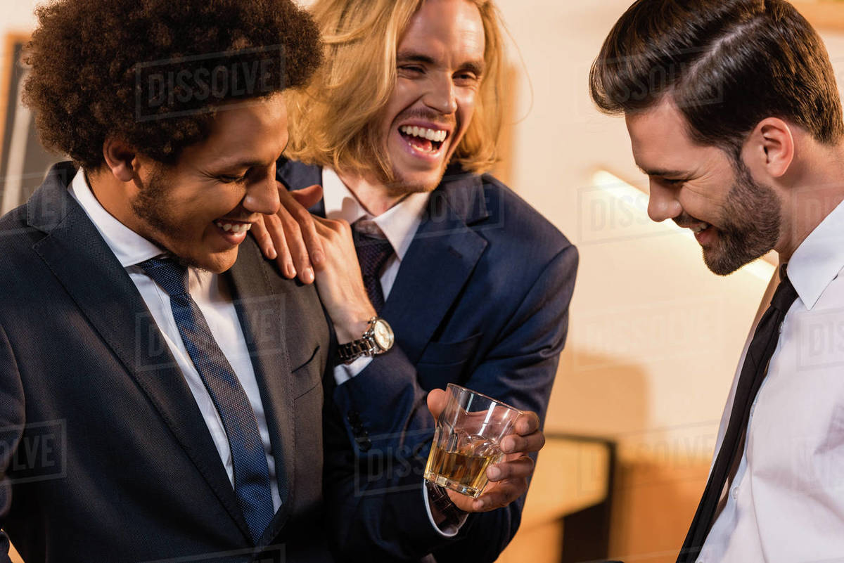 Three multiethnic businessmen laughing while drinking whiskey in bar ...