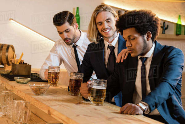 Young multiethnic men in formal wear drinking beer and talking in bar ...