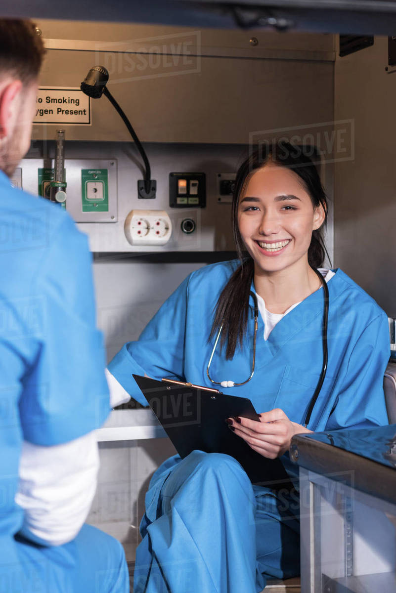 Smiling Asian paramedic sitting in ambulance and looking at camera ...