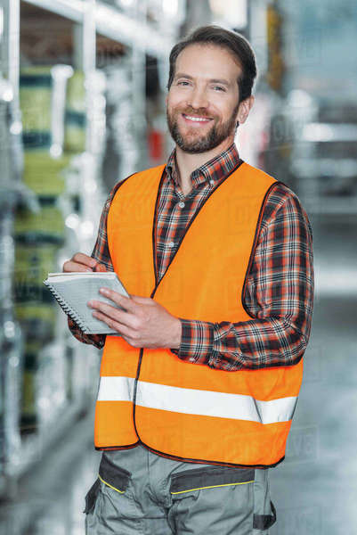 Smiling worker in safety vest writing in notepad in storehouse ...